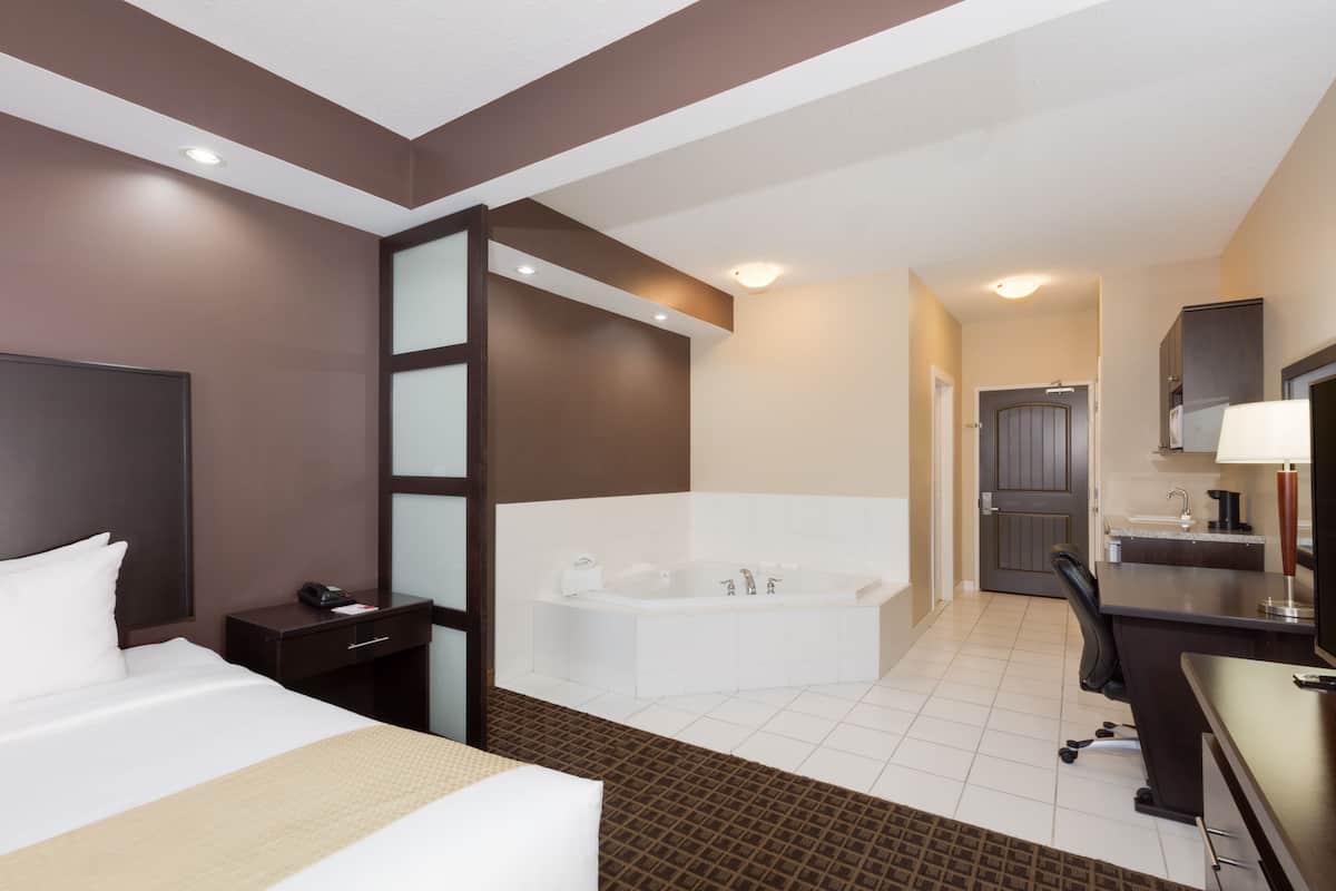 Quarto de hotel com uma cama grande, banheiro separado com banheira, escrivaninha com cadeira e uma pequena luminária de mesa.