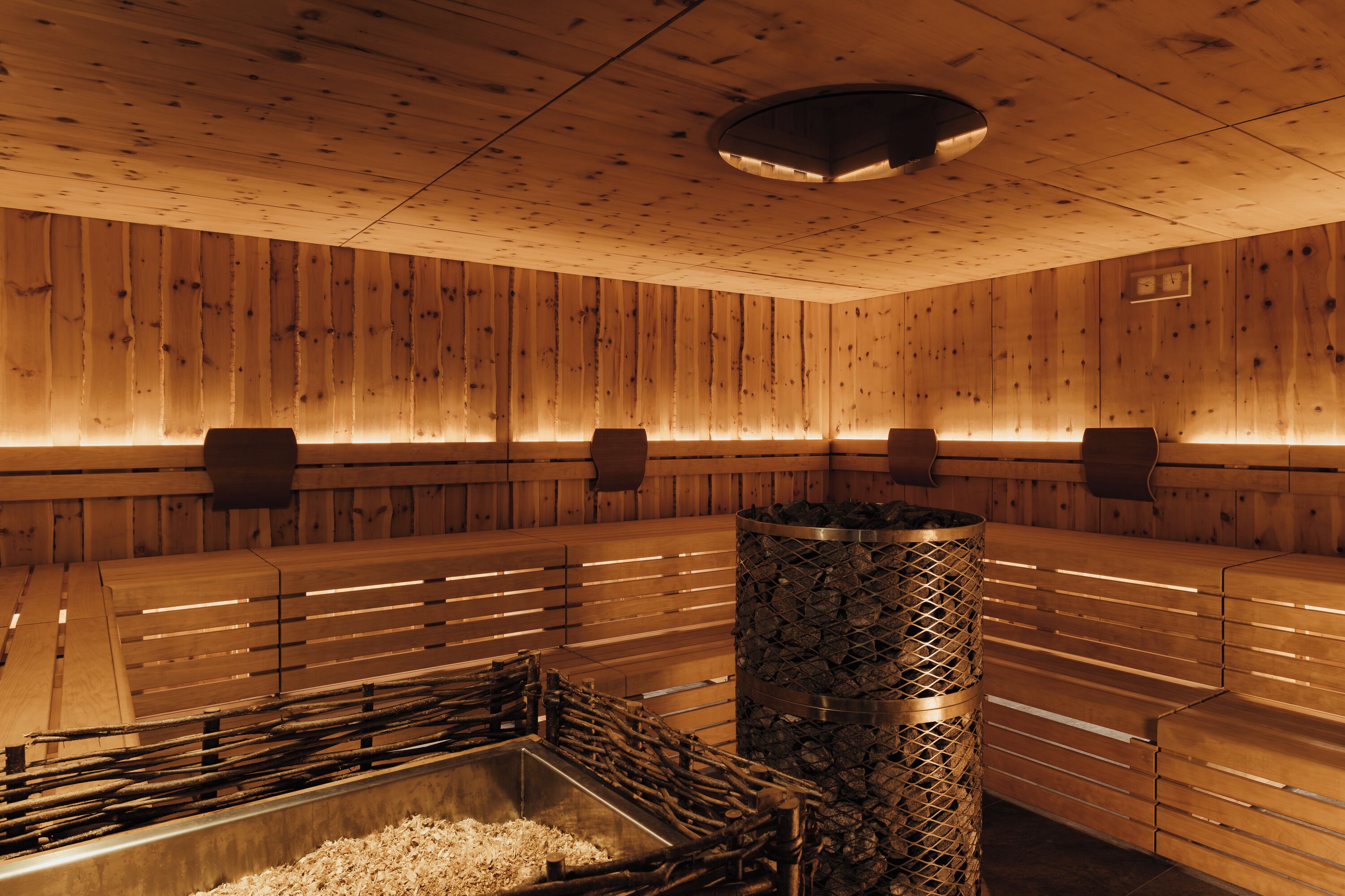 Uma sauna com paredes e bancos de madeira, uma lareira central e uma luminária no teto.