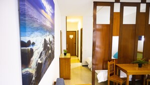 Quarto de hotel com divisória de madeira, uma mesa com cadeira, uma pequena mesa com uma planta e uma cama com cobertor branco.