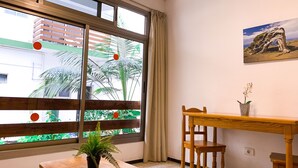 Quarto com uma grande janela, uma mesa de madeira, uma cadeira, um vaso com planta e um quadro na parede.
