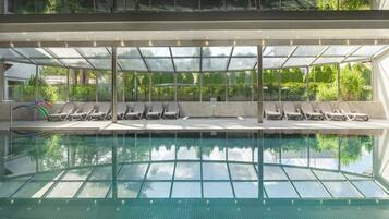 Piscina interior moderna com paredes de vidro transparente, cadeiras de descanso e vista para áreas verdes.