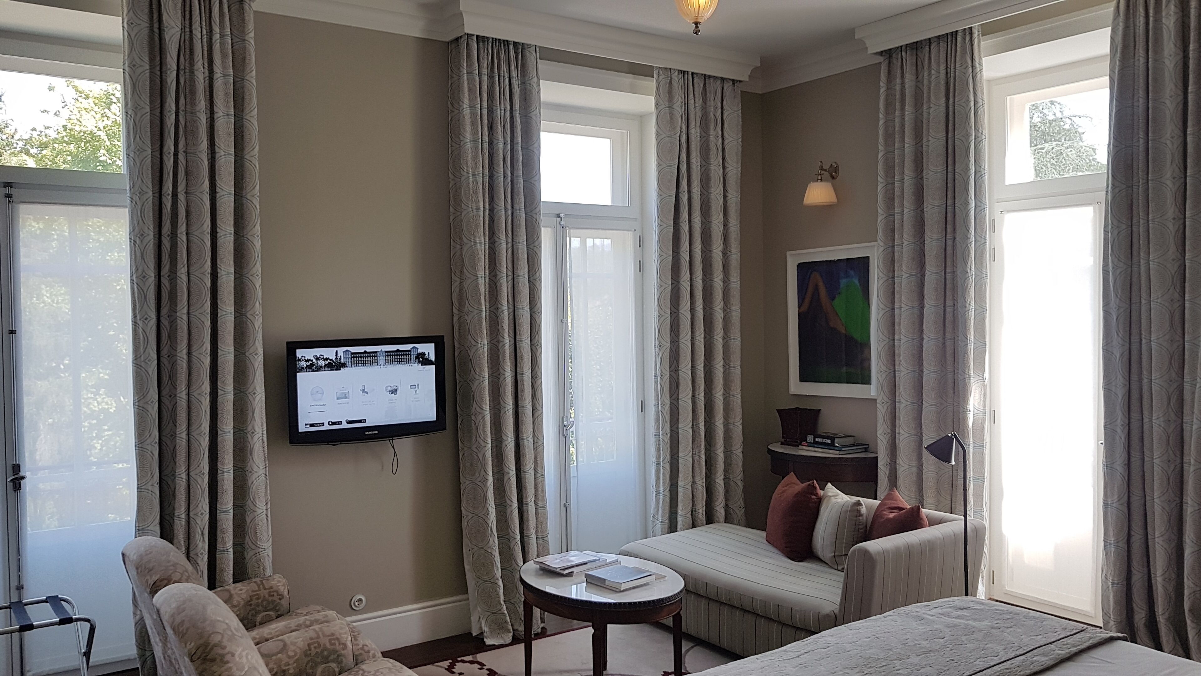 Quarto de hotel com cama, sofá, mesa pequena, televisão fixada na parede e janela com cortinas.