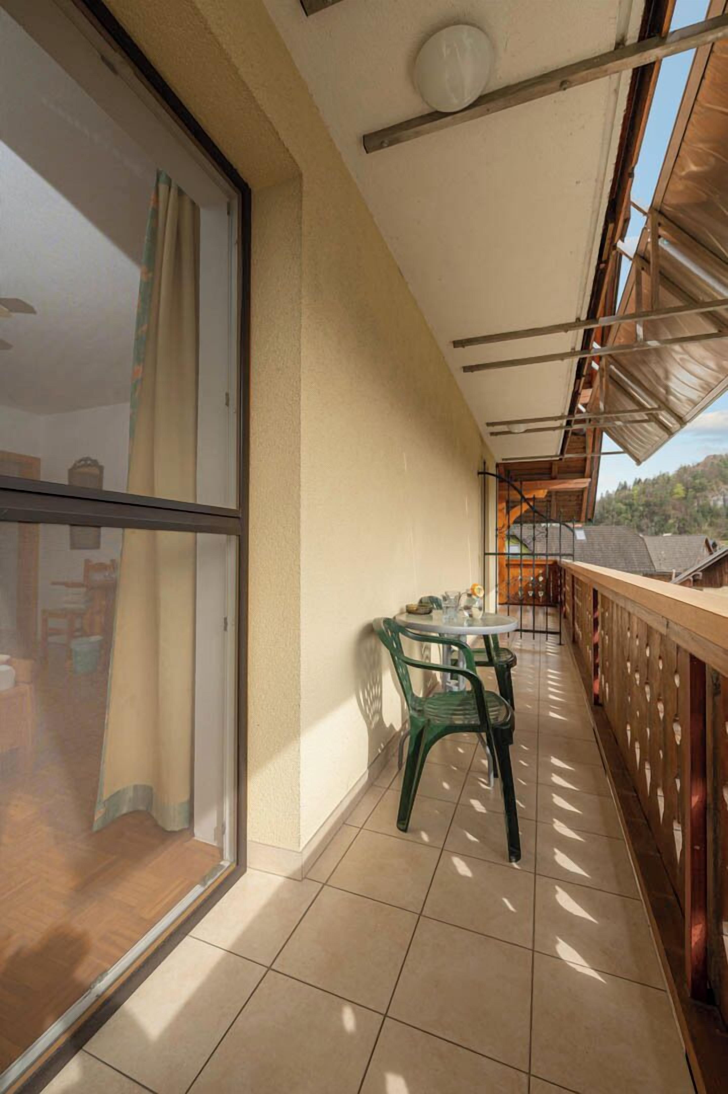 Uma varanda com porta de vidro, uma mesa com cadeiras e vista para um edifício com telhado inclinado.