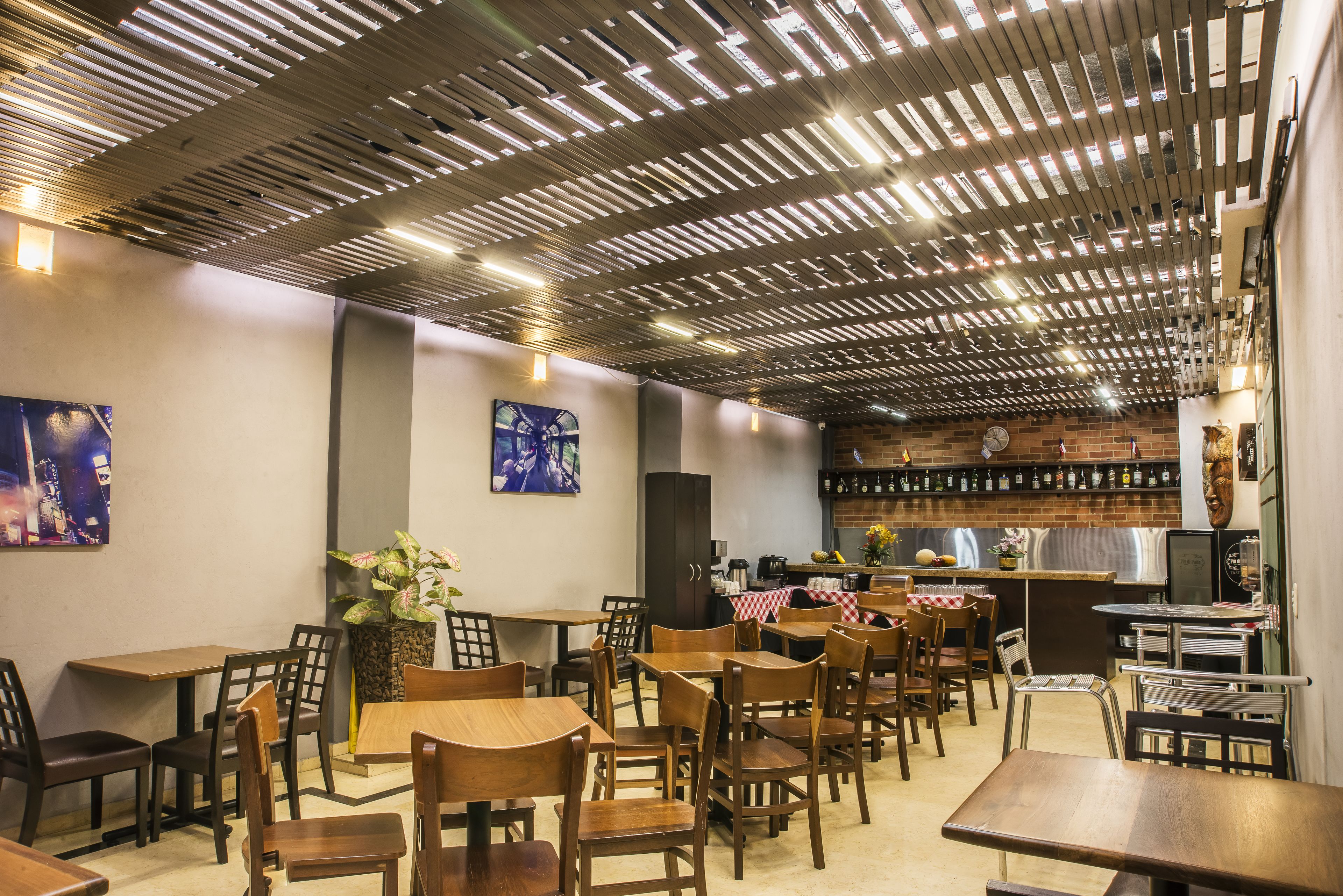 Um restaurante com mesas e cadeiras de madeira, uma parede de tijolos e teto com iluminação linear.