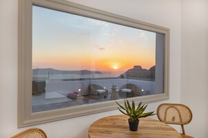 Quarto com grande janela com vista para o pôr do sol, mesa redonda de madeira com planta em vaso e cadeiras de vime.