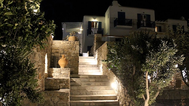 Uma escadaria de pedra leva a um prédio branco com persianas azuis, cercado por vegetação e iluminado por iluminação ambiente.