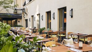 Uma área de refeições ao ar livre com várias mesas e cadeiras de madeira, rodeada por plantas em vasos e equipada com copos de vinho e guardanapos.