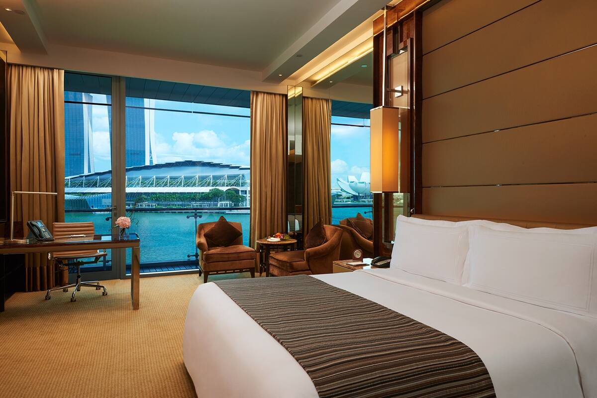 Quarto de hotel com uma cama grande, uma mesa com telefone, uma cadeira e vista para uma ponte e para a água.