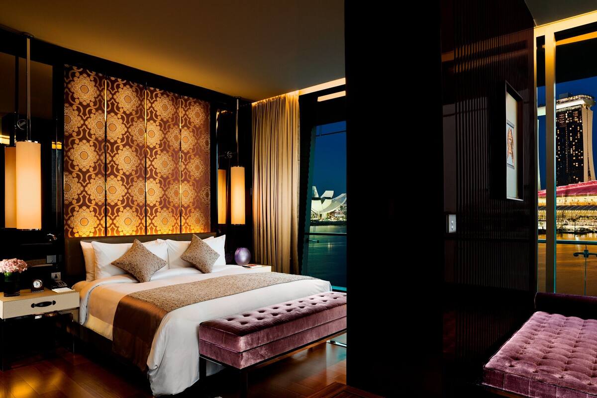 Quarto de hotel moderno com uma cama grande, um banco e vista para a paisagem urbana à noite.