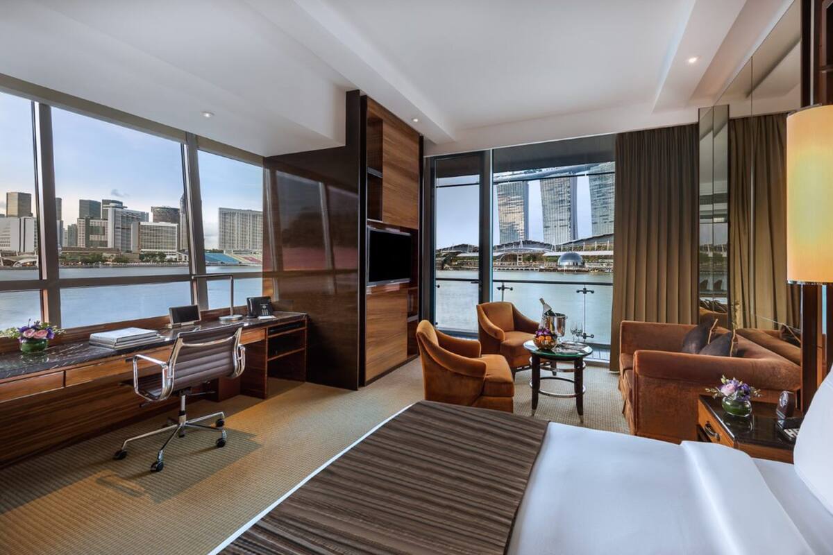 Quarto de hotel com uma cama grande, uma escrivaninha com cadeira, um sofá e vista para o horizonte urbano através de amplas janelas.