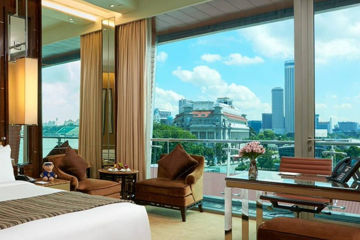 Quarto de hotel com uma cama grande, duas poltronas, uma mesa pequena e vista para a paisagem urbana.