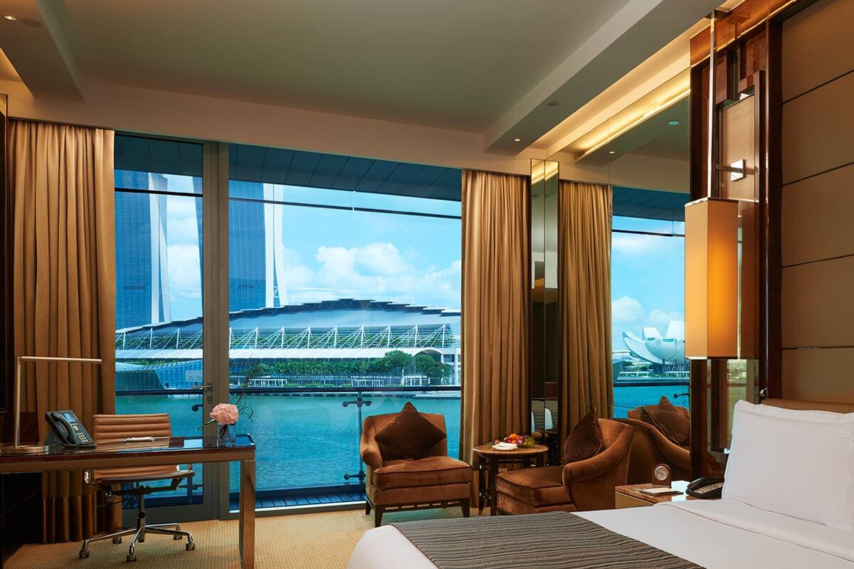Quarto de hotel com uma grande janela que oferece vista para uma ponte e para a água. O quarto tem uma cama, duas poltronas, uma escrivaninha com abajur e uma pequena mesa com um vaso de flores.
