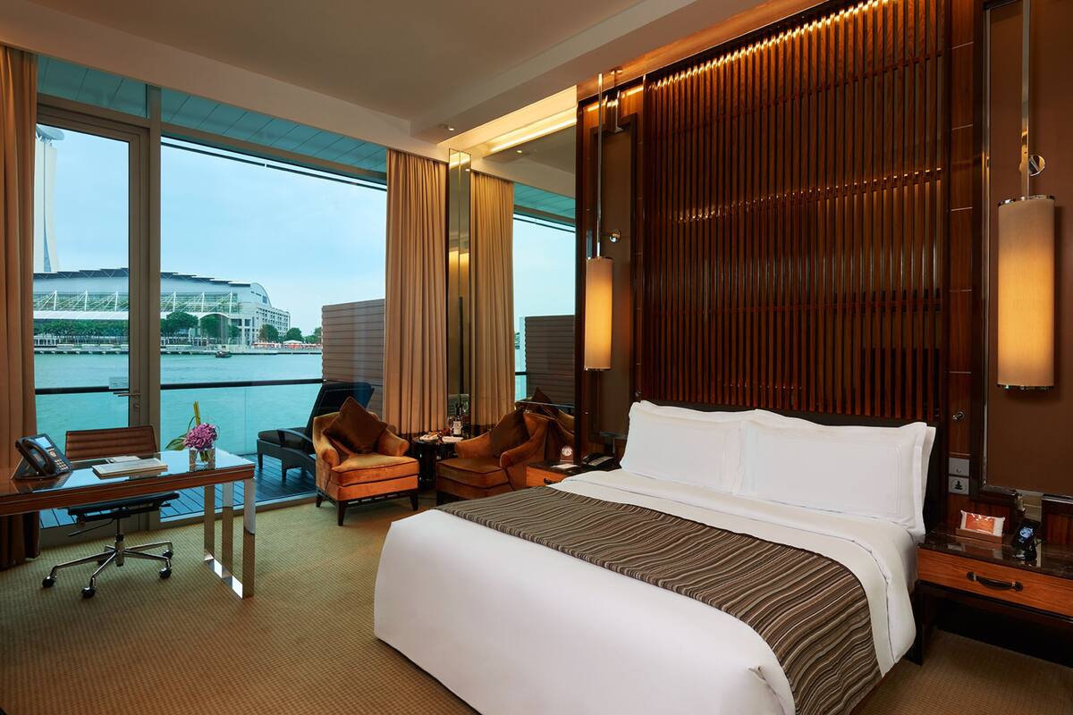 Quarto de hotel com uma cama grande, uma escrivaninha com telefone, duas poltronas e vista para um rio e uma ponte.