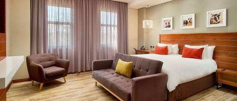 Quarto de hotel com cama grande, sofá, poltrona e escrivaninha. Três quadros decoram a parede.