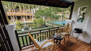 Varanda com cadeiras de vime e uma pequena mesa, com vista para uma piscina e vegetação exuberante.