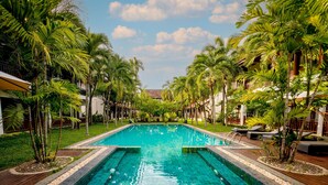 Uma piscina retangular cercada por palmeiras e um deck de madeira, com um edifício ao fundo.