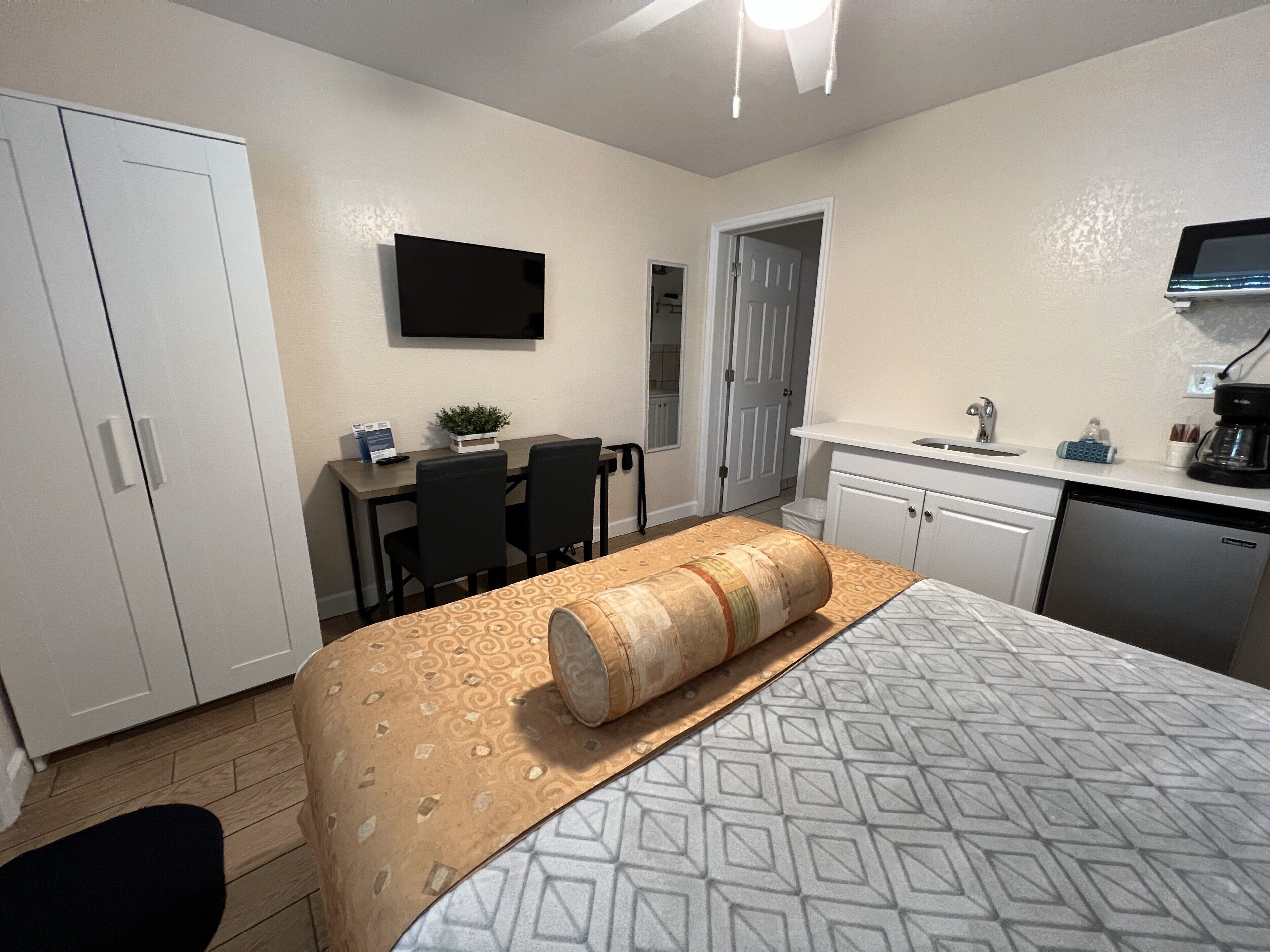 Quarto compacto com uma cama, escrivaninha, cadeira, televisão, uma pequena planta e cozinha compacta.