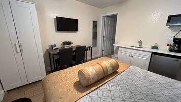 Quarto compacto com uma cama, escrivaninha, cadeira, televisão, uma pequena planta e cozinha compacta.