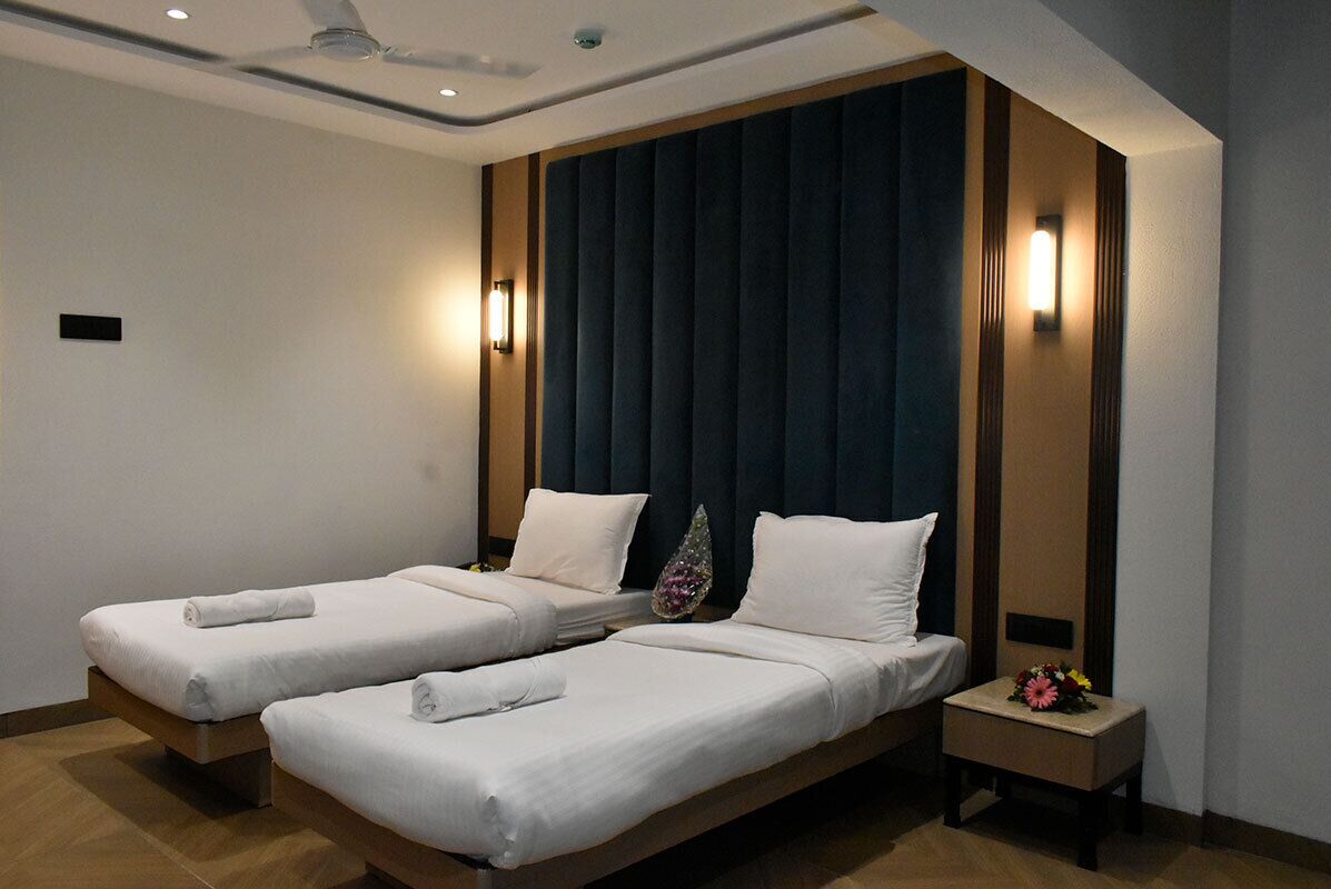 Quarto de hotel com duas camas de solteiro, cabeceira escura, abajures e uma pequena mesa com um arranjo de flores.
