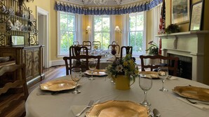 Uma sala de jantar com uma mesa grande posta para refeição, rodeada por cadeiras, um candelabro e uma lareira.