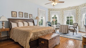 Quarto com cama de madeira, mesa de cabeceira, cômoda, ventilador de teto e janela com cortinas.