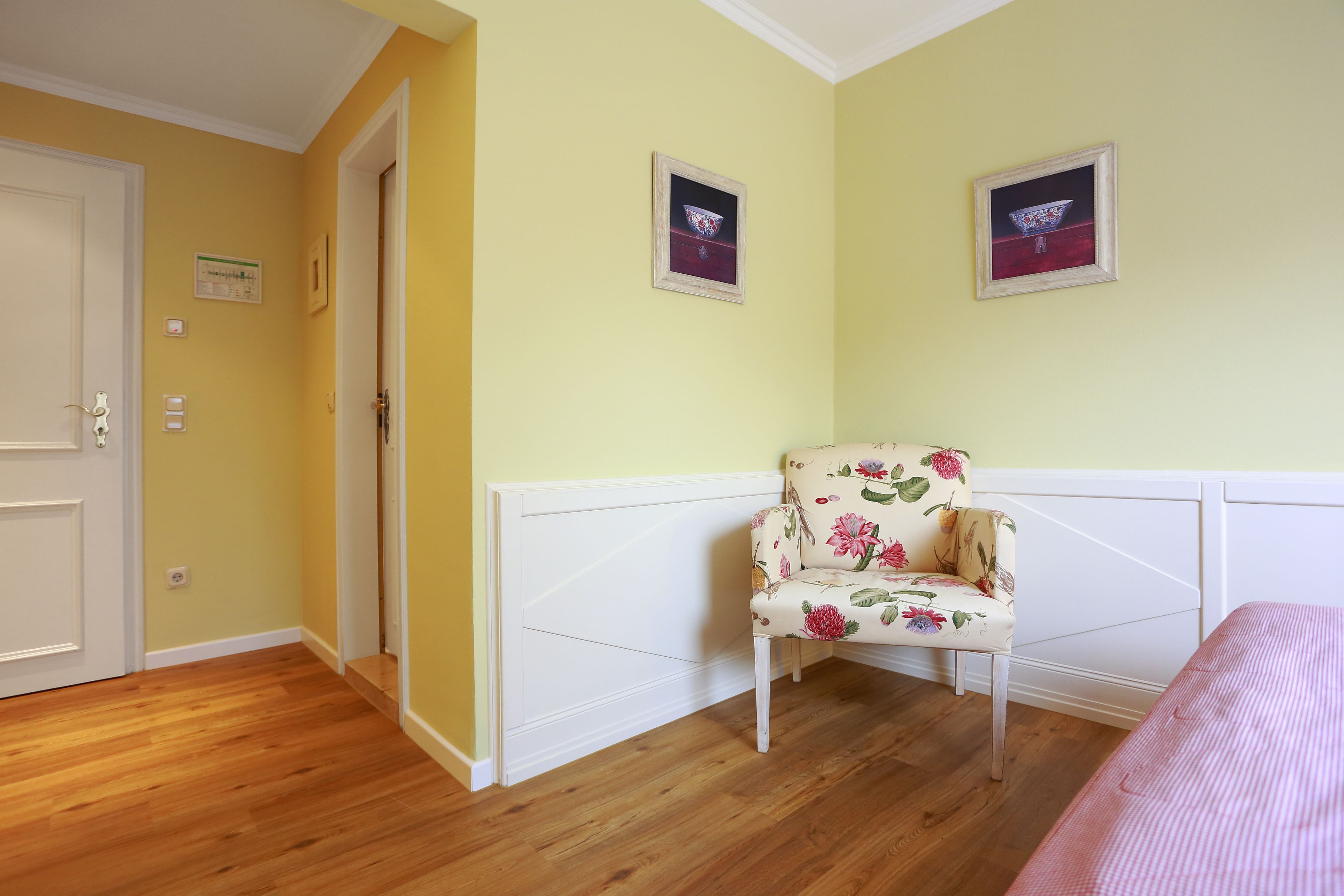 Quarto com piso de madeira, poltrona estampada, uma cama e duas fotos emolduradas na parede.