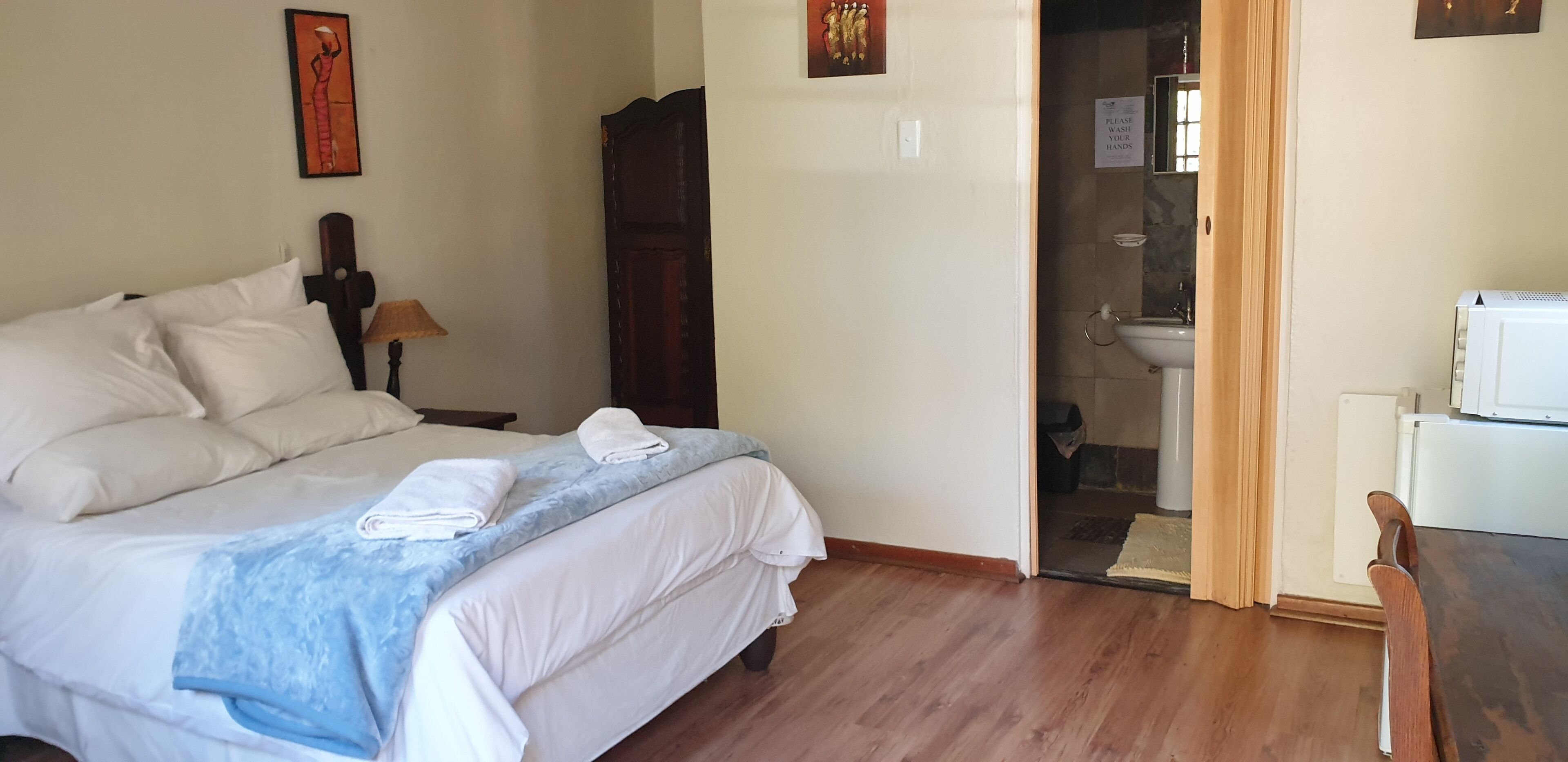 Quarto com cama, mesa de cabeceira, luminária, porta de madeira e banheiro visível por uma porta aberta.