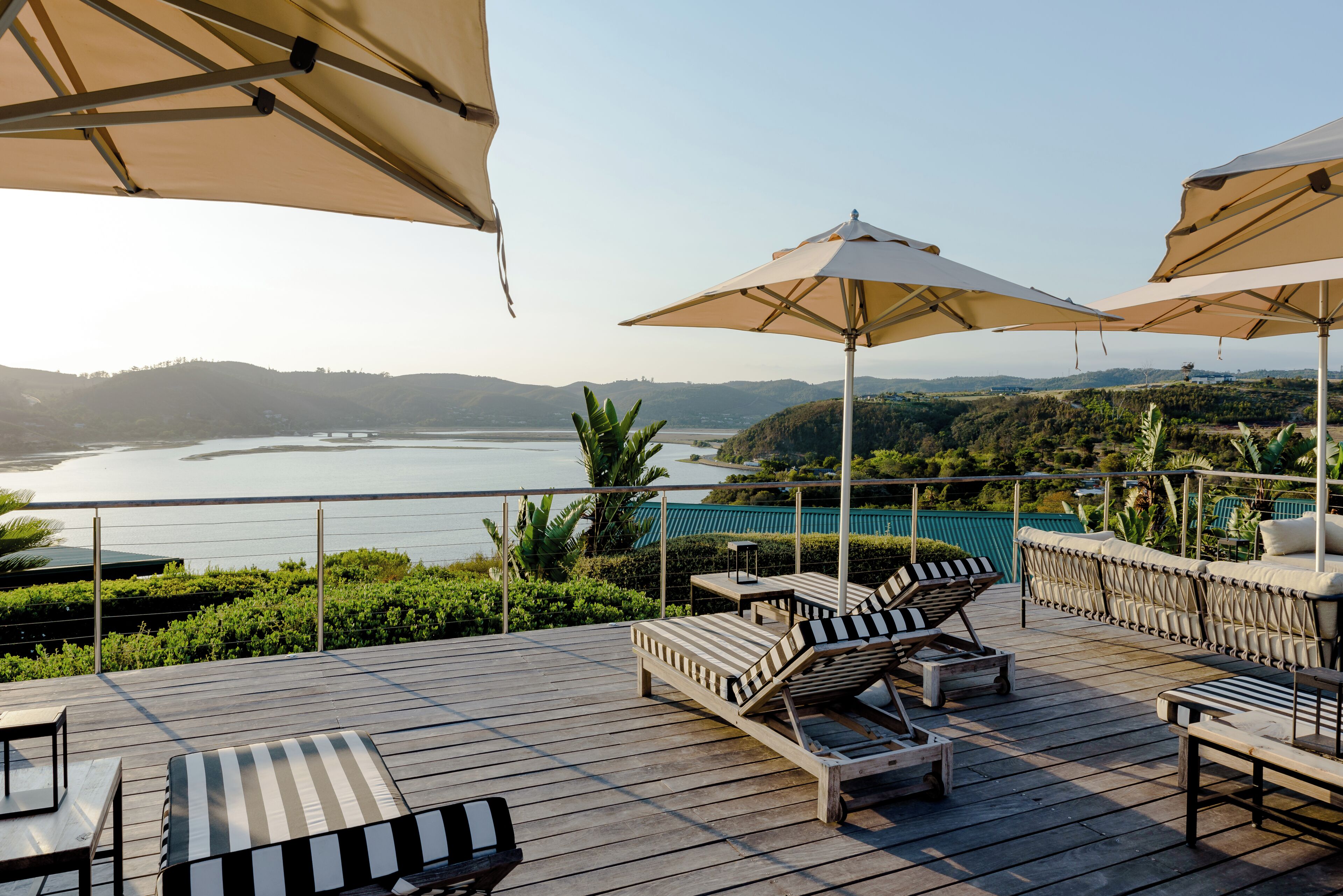 Um deck com cadeiras de lounge e guarda-sóis com vista para um corpo de água e colinas.
