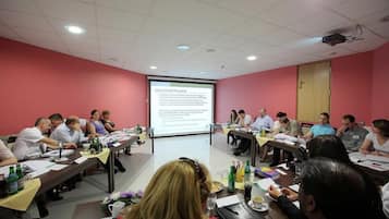 Sala de conferência com tela de projeção, mesas e participantes.