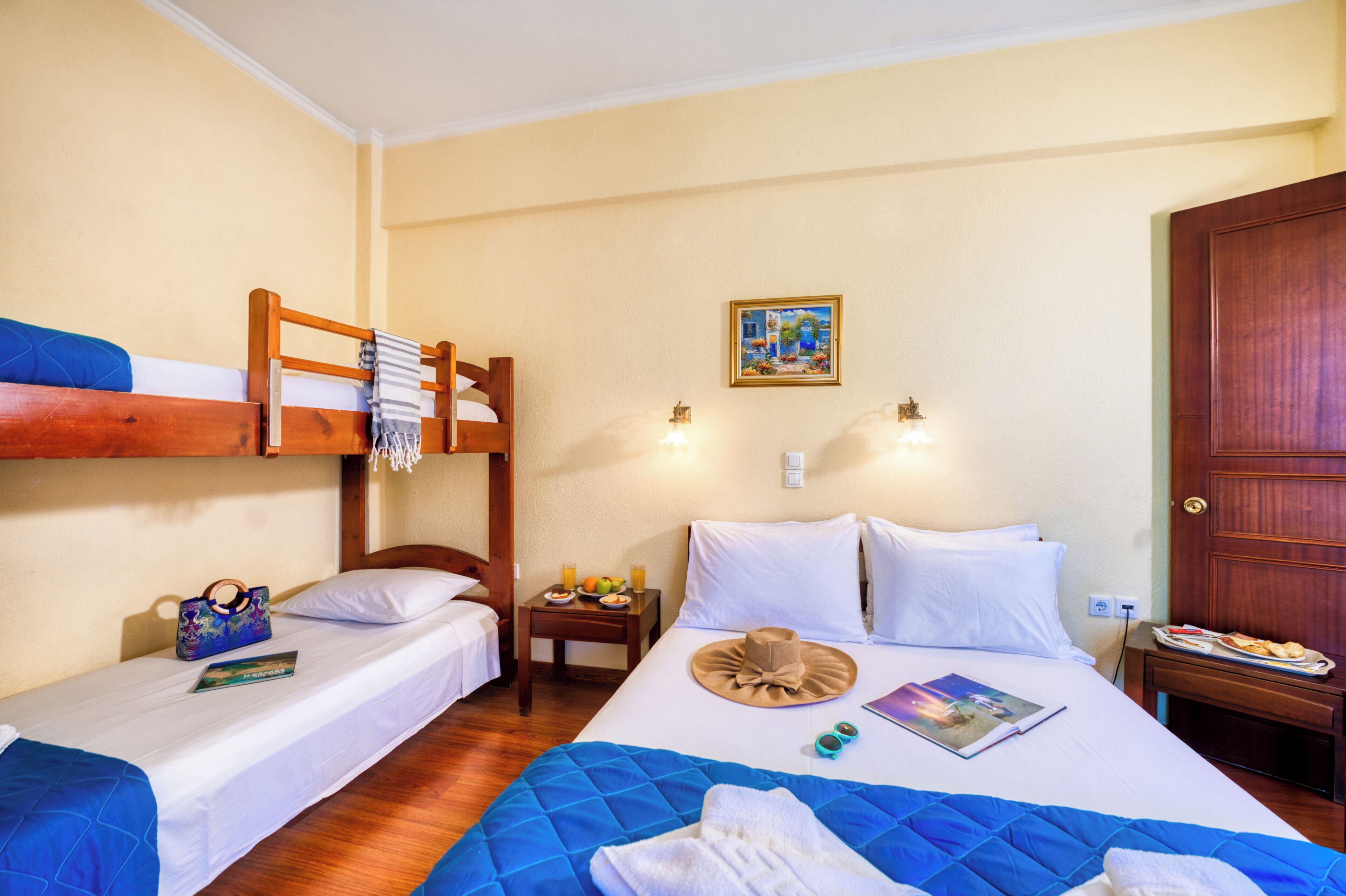 Quarto de hotel com beliche, uma cama de solteiro, mesa de cabeceira com uma fruteira, luminária na parede e uma porta.