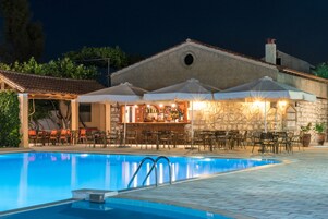 Uma piscina com espreguiçadeiras e guarda-sóis, área de bar com cadeiras e um edifício de pedra com telhado de azulejos.