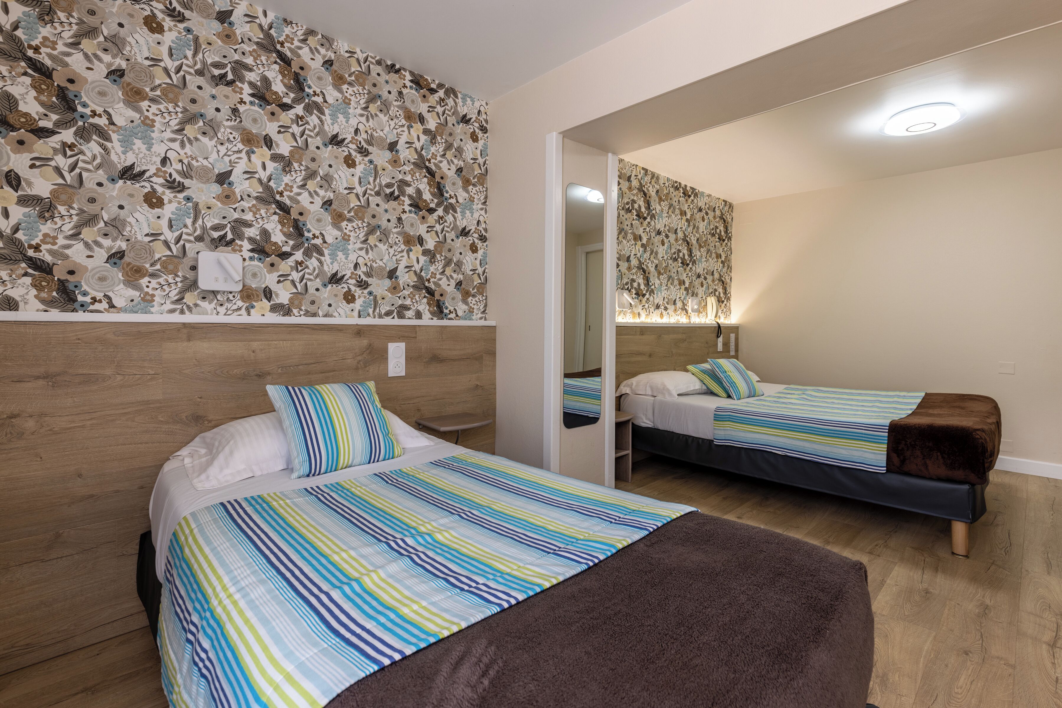 Quarto com duas camas, cabeceira de madeira, parede com padrão floral e um espelho refletindo o ambiente.