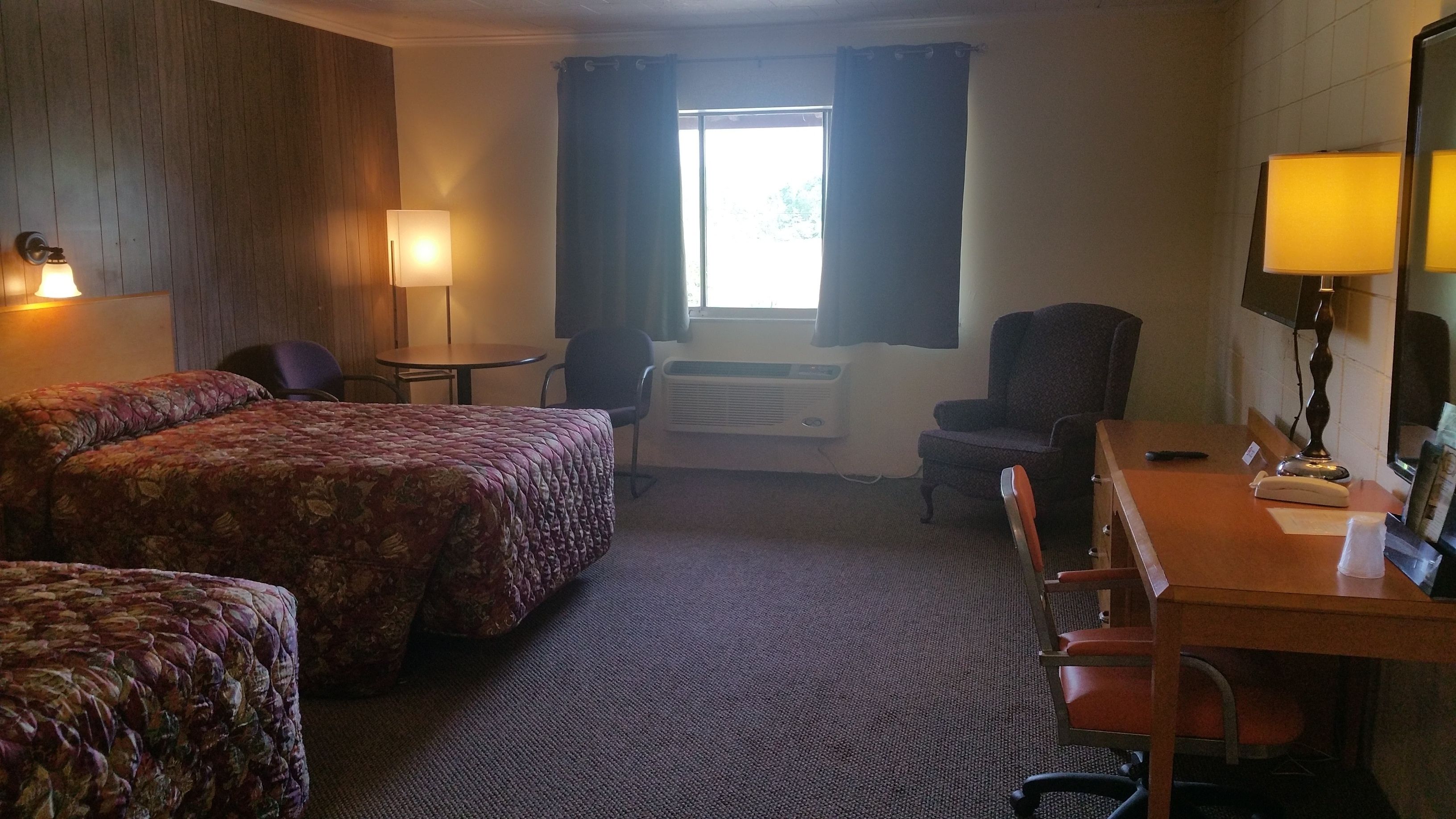 Quarto de hotel com duas camas, uma mesa, uma cadeira, uma luminária e uma janela com cortinas.