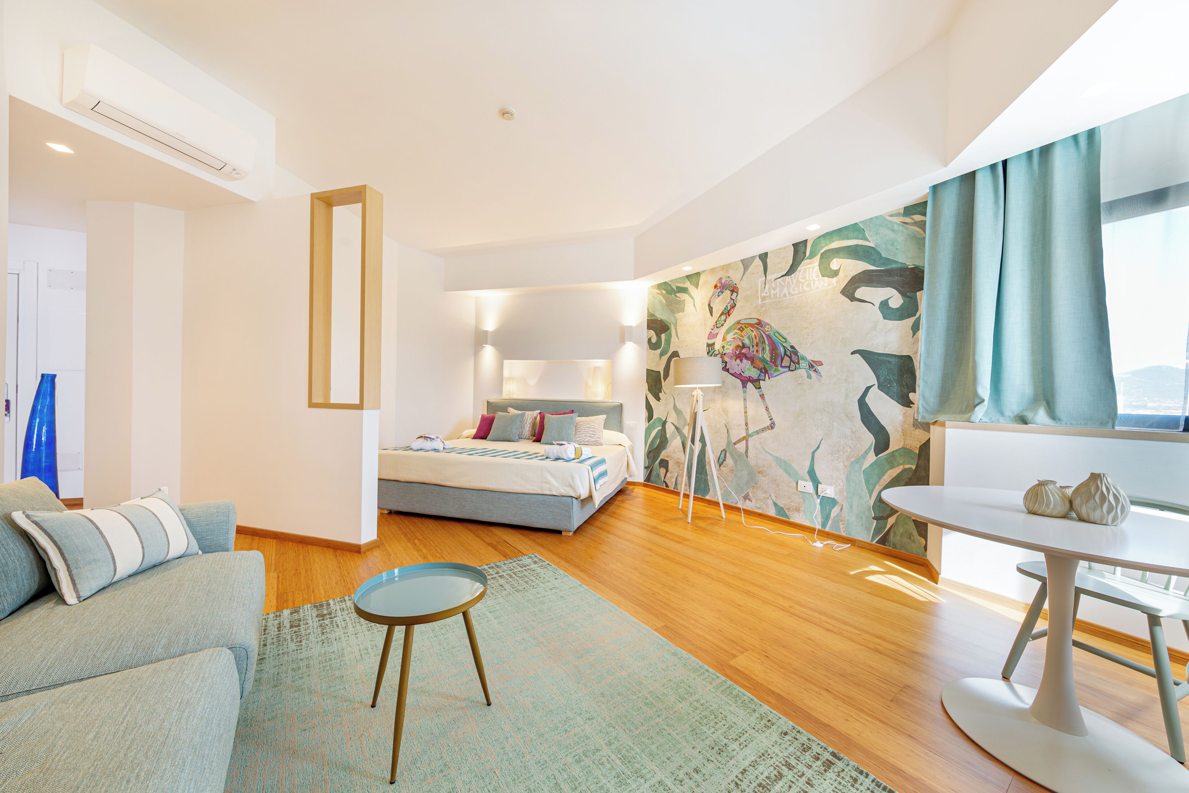 Quarto de hotel moderno com cama, sofá, mesa de jantar e uma janela grande.