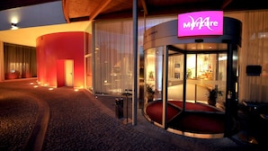 Uma entrada de hotel moderna com porta de vidro, fachada iluminada em vermelho e uma área de recepção visível no interior.