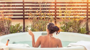 Uma pessoa relaxando em uma banheira de hidromassagem com um deck de madeira e plantas ao fundo.