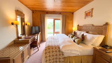Quarto de hotel com teto de madeira, cama com roupa de cama estampada, televisão sobre uma escrivaninha de madeira e vista para o verde lá fora.