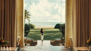 Quarto com uma grande janela com vista para uma praia, o oceano e um jardim tropical.
