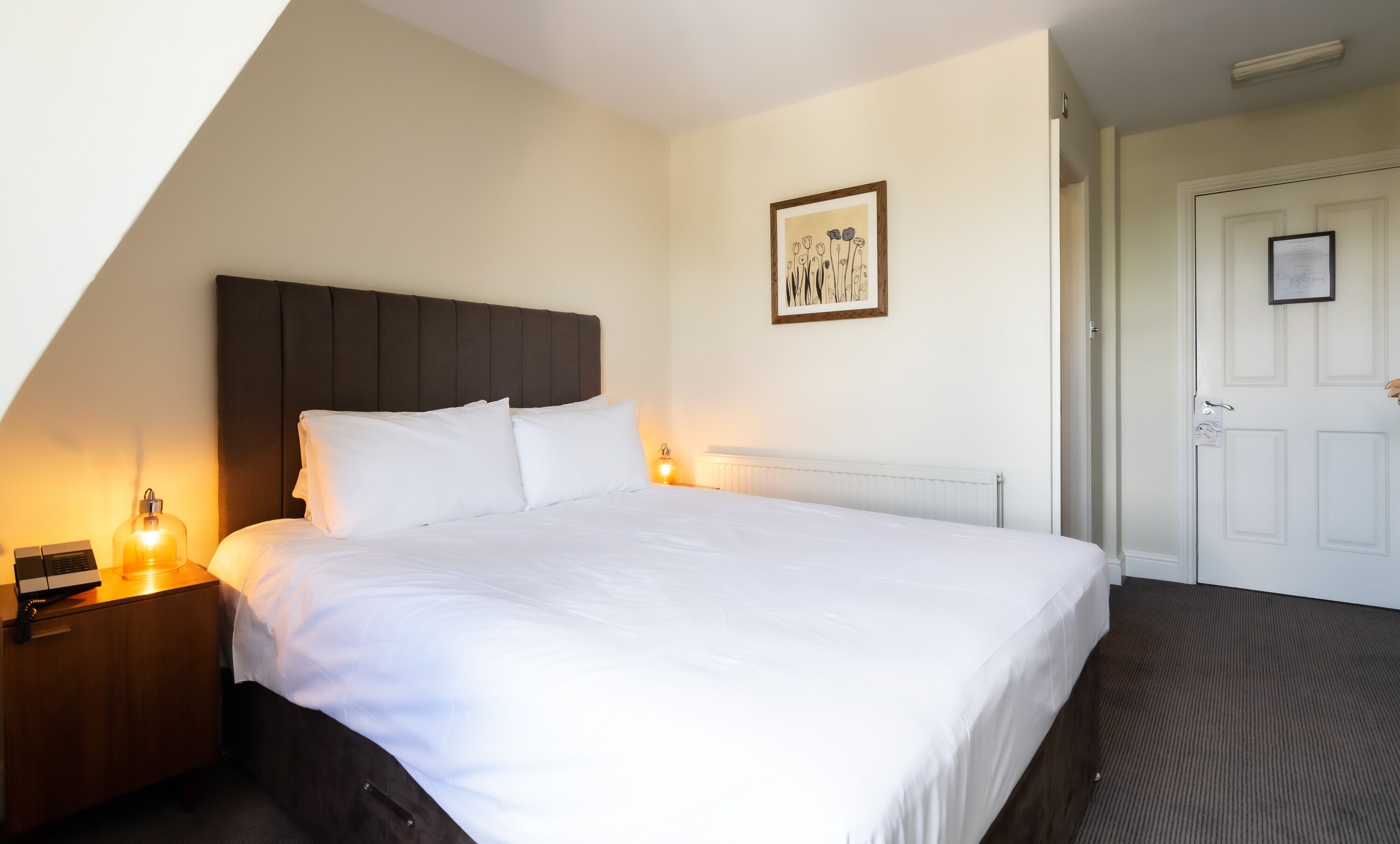 Quarto de hotel com uma cama grande, mesa de cabeceira com abajur, quadro na parede e porta com chave na fechadura.