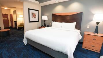 Quarto de hotel com uma cama grande, uma mesa de cabeceira com luminária, uma cadeira e uma escrivaninha com computador.
