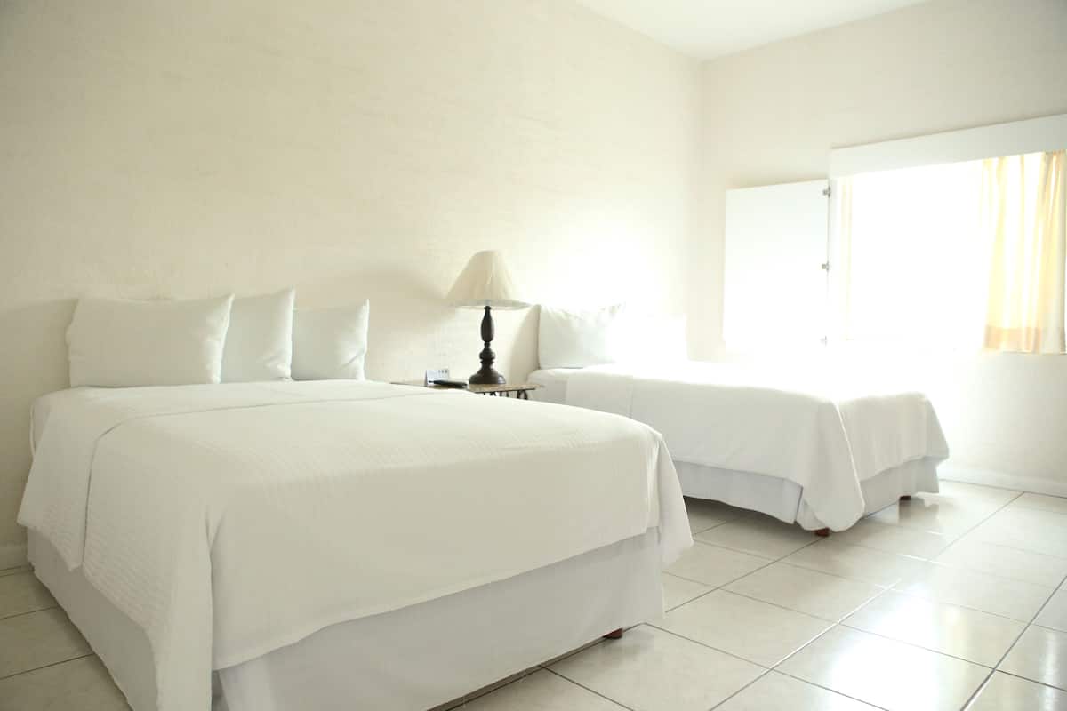 Quarto de hotel com duas camas, uma mesinha de cabeceira, uma luminária e uma janela com cortinas.