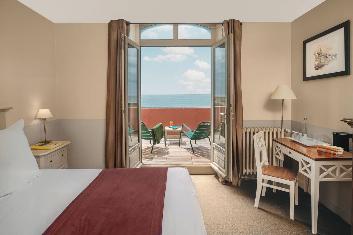 Quarto de hotel com cama, escrivaninha, cadeira, mesa e varanda com vista para o mar.