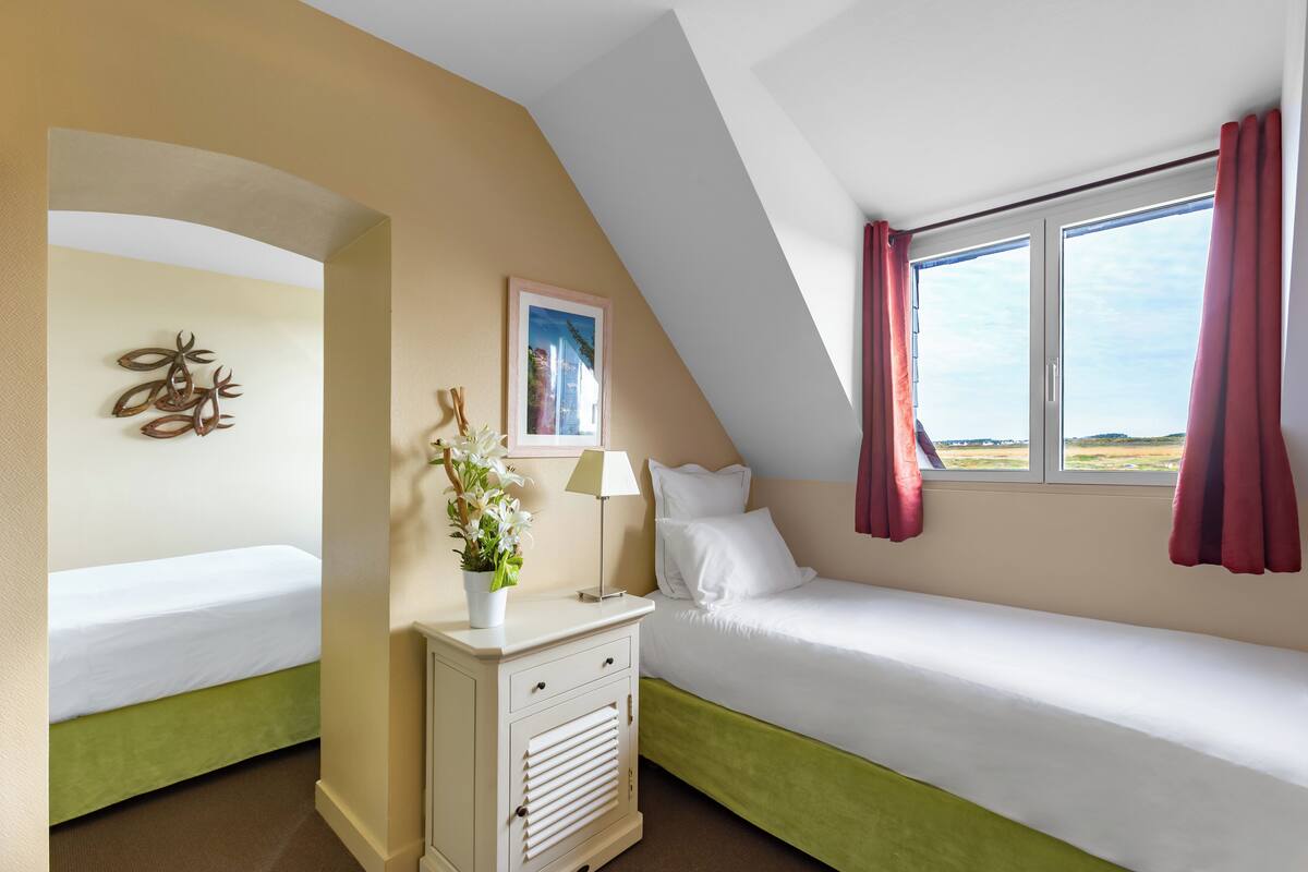 Quarto com cama, mesa de cabeceira, abajur, quadro emoldurado e janela com cortinas.