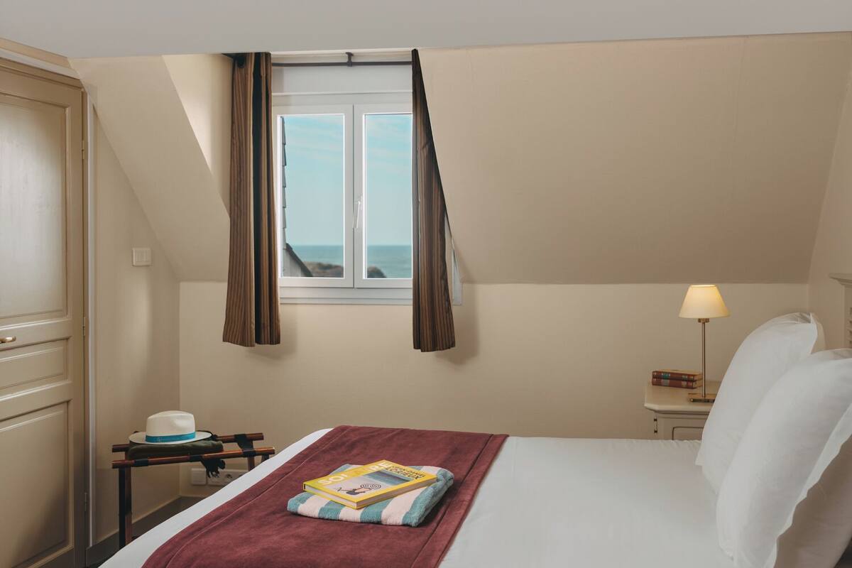 Quarto de hotel com cama, janela com cortinas, mesa de cabeceira com abajur e vista para o mar.