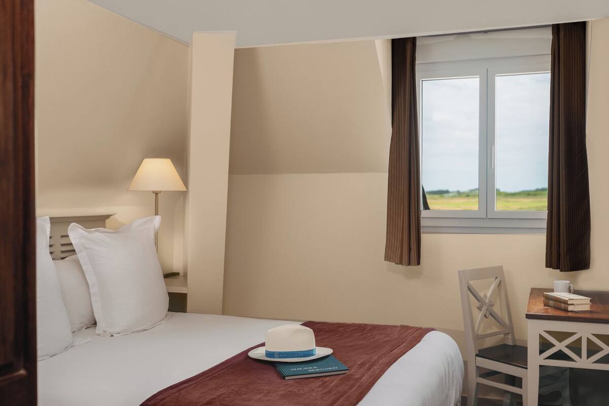 Quarto de hotel com cama, escrivaninha, cadeira, luminária, janela com cortinas e vista para o exterior.