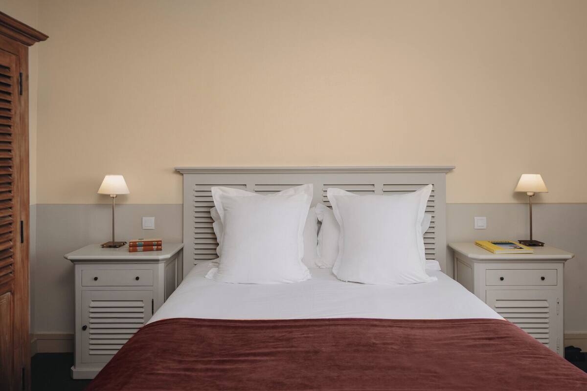 Cama bem arrumada com lençóis brancos e manta marrom, com duas mesas de cabeceira com luminárias e livros de cada lado.