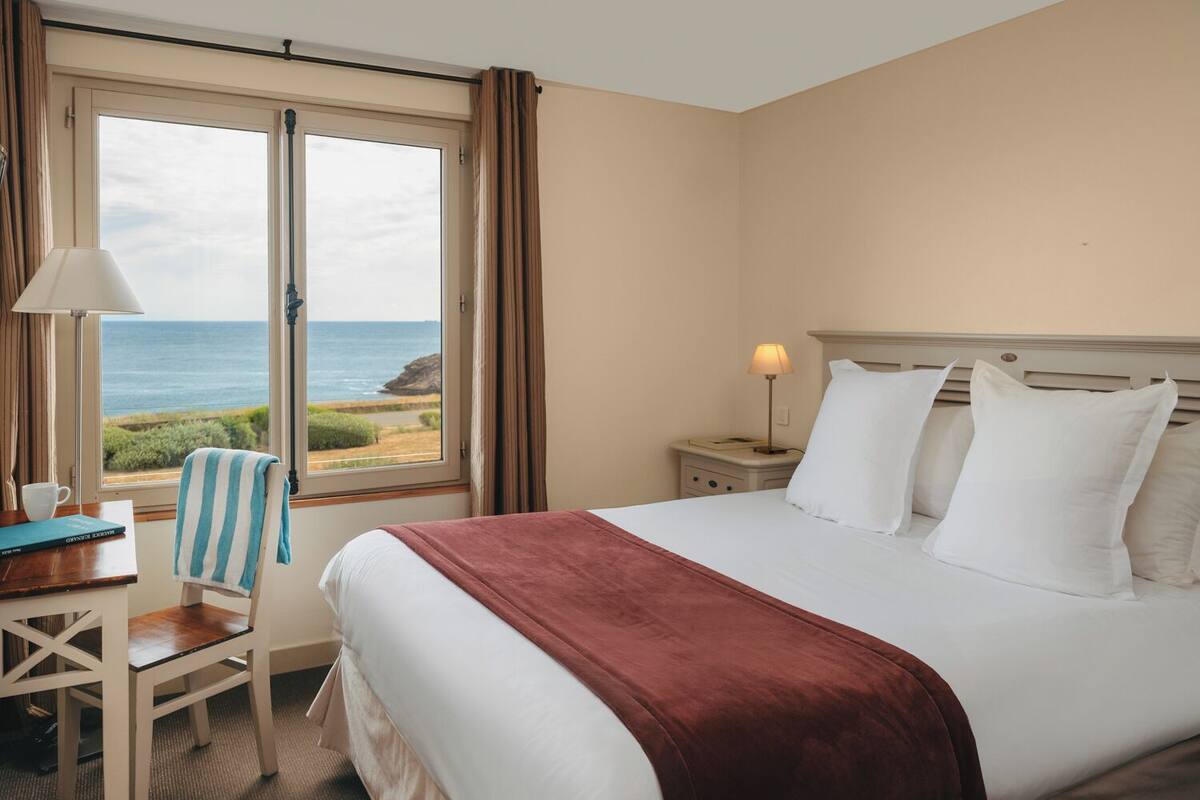 Quarto de hotel com cama, uma escrivaninha com luminária, uma cadeira com toalha e vista para o mar através de uma janela grande.
