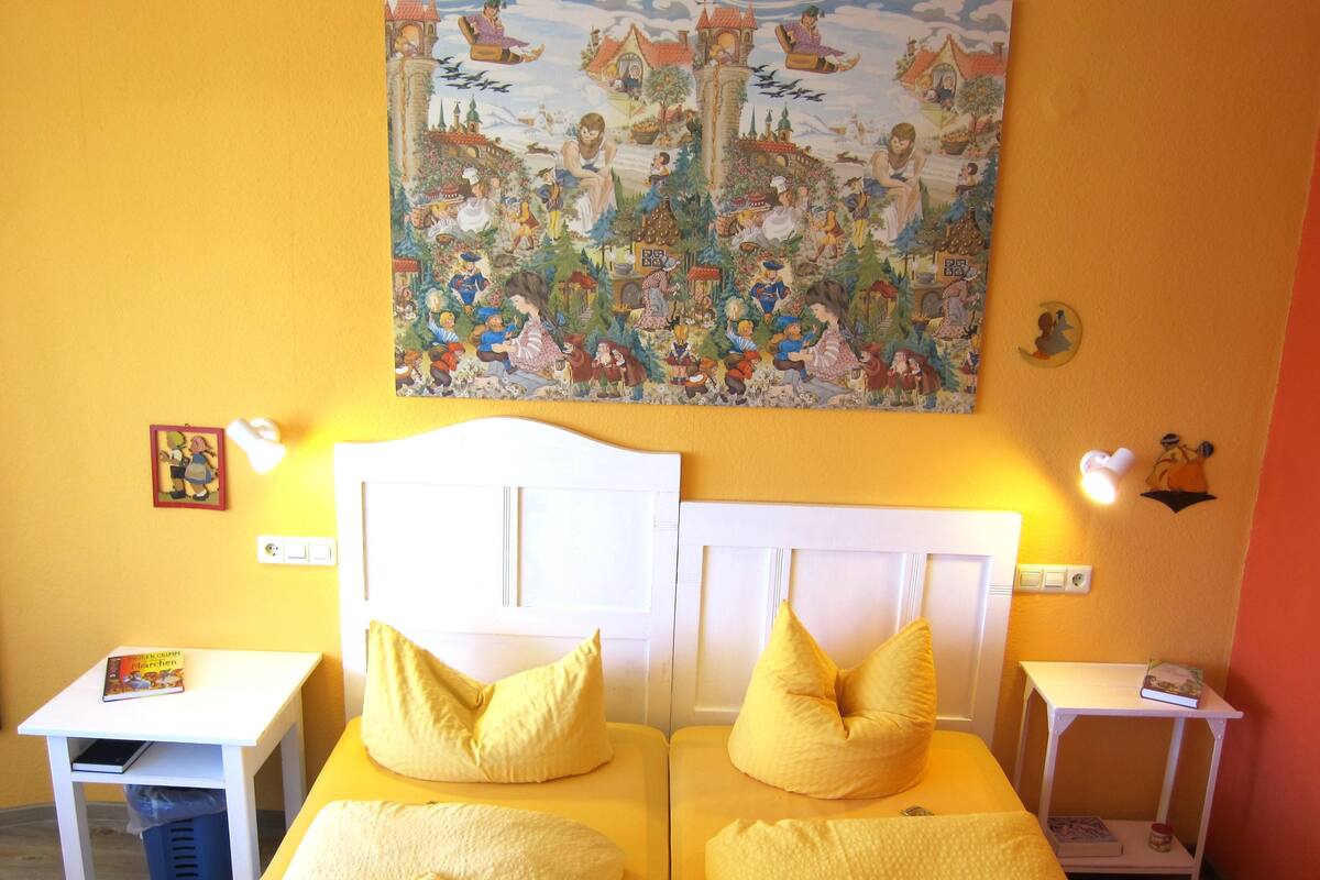 Quarto com duas camas, um mural na parede e uma pequena mesa com um livro.
