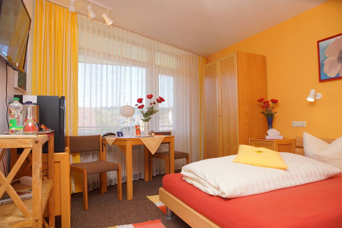 Quarto de hotel com cama, escrivaninha, cadeira, mesa com vaso de flores e televisão.