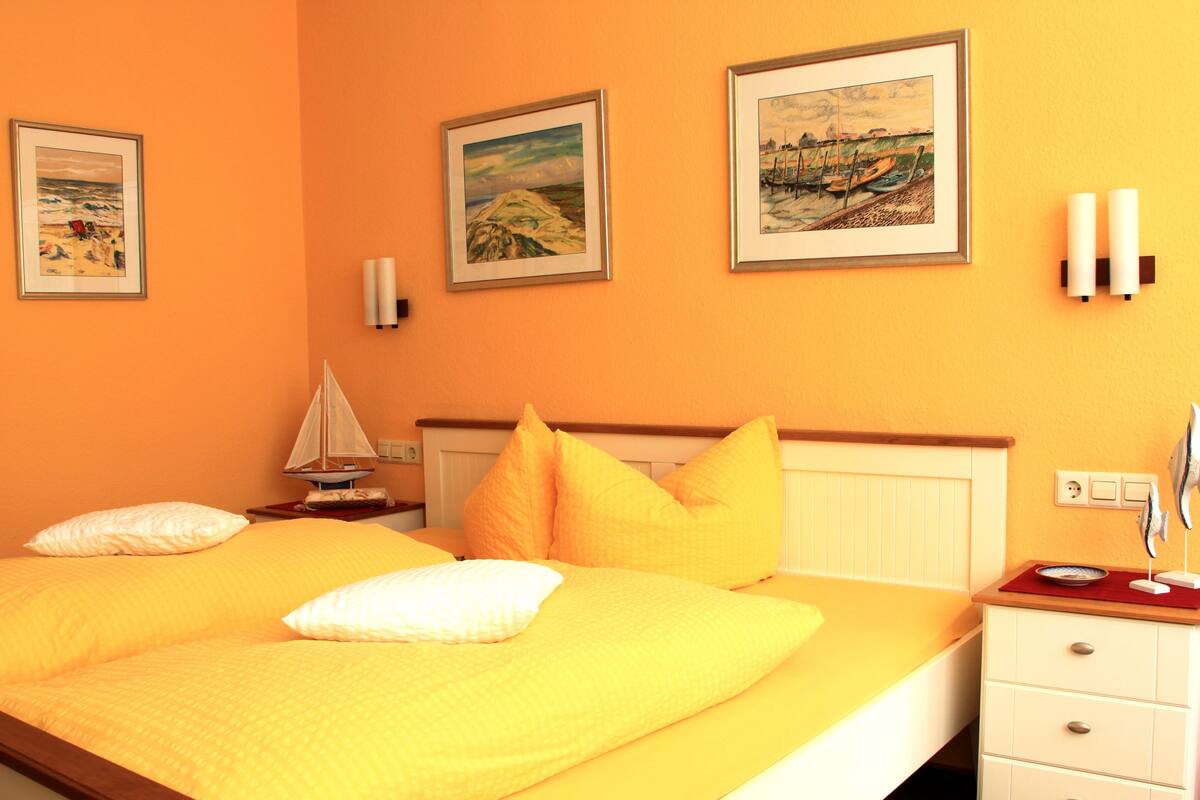 Quarto de hotel com uma cama grande, duas pinturas na parede, uma mesa de cabeceira com abajur e um armário com gavetas.